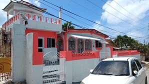 Uma casa de dois andares com fachada vermelha e branca, entrada protegida por portão e um carro branco estacionado em frente.