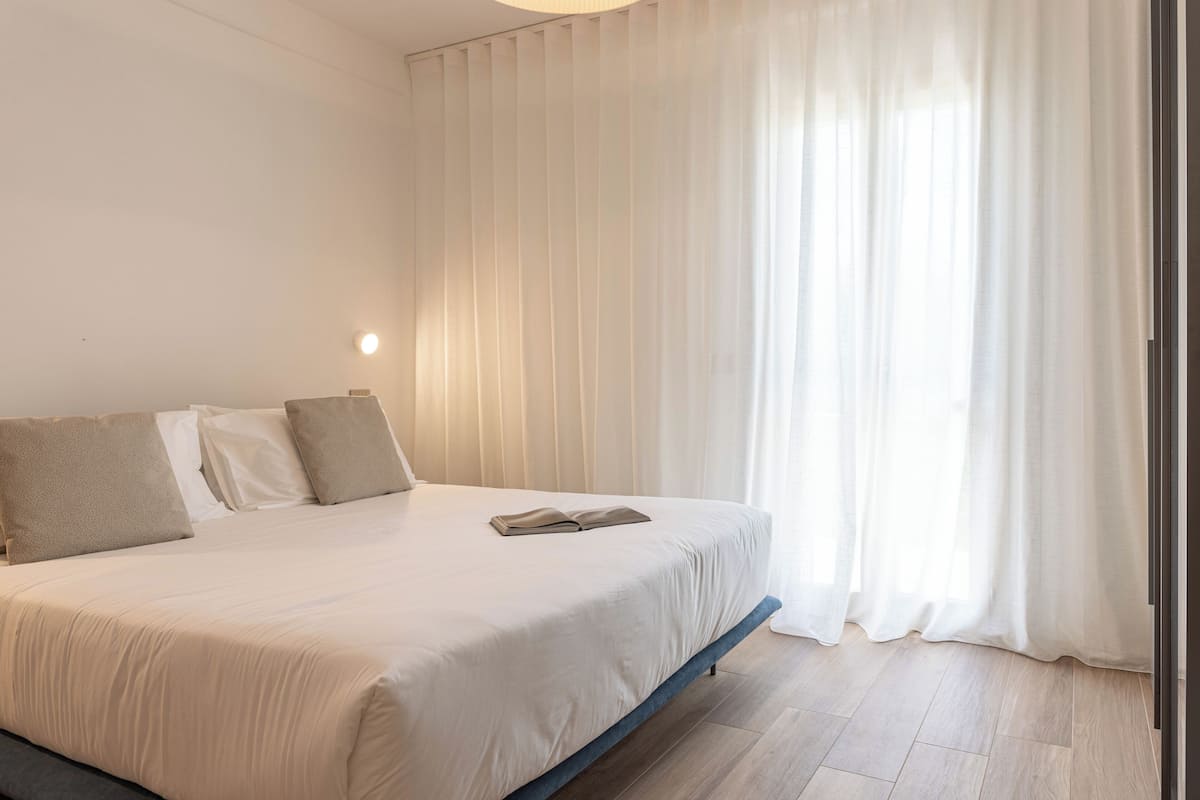 Quarto com cama, almofadas, um livro e cortinas finas.