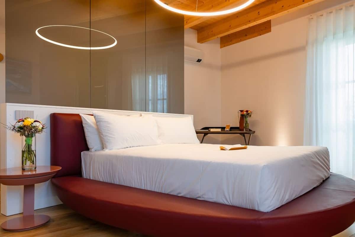 Quarto moderno com cama curva, mesa de cabeceira com um vaso e um espelho grande.