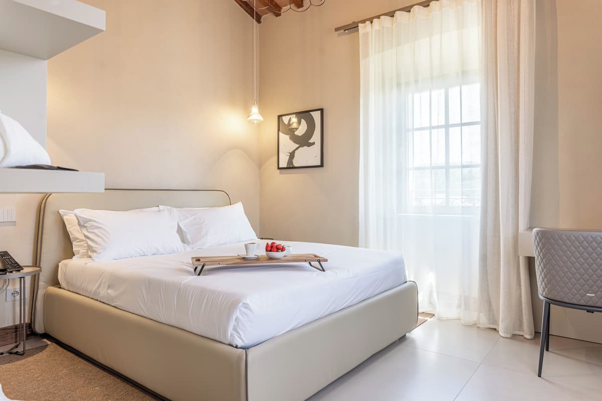 Quarto de hotel moderno com cama, mesa de cabeceira com abajur, cadeira e janela com cortinas.