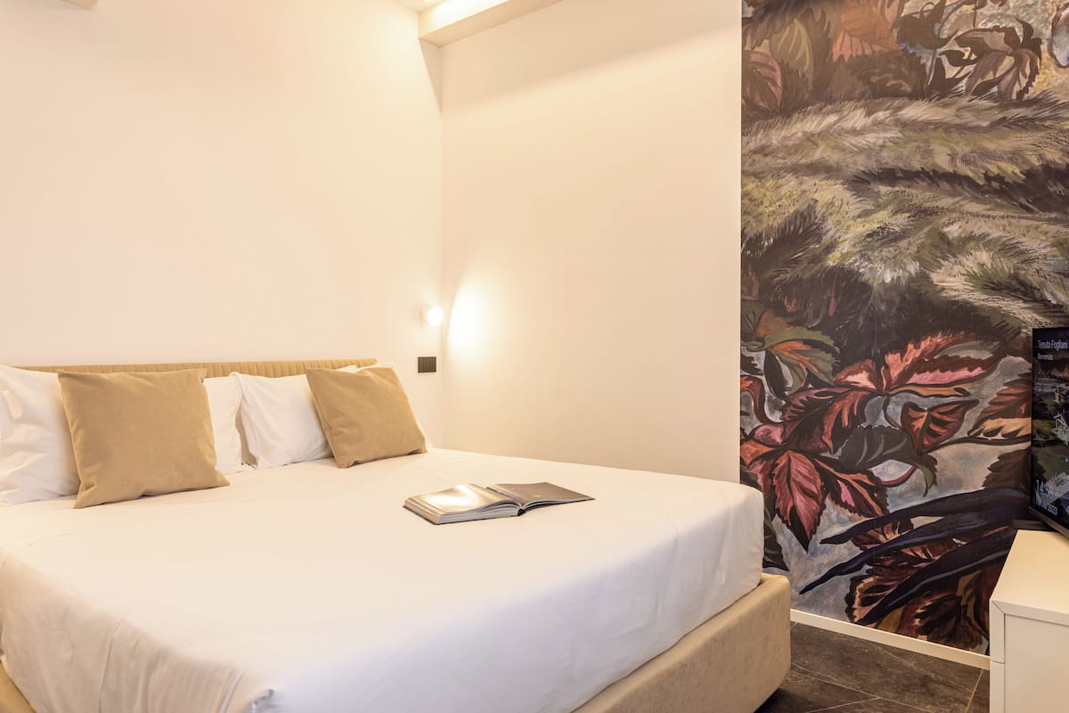 Uma cama bem arrumada com roupa de cama branca e bege, um livro sobre ela e um mural na parede com tema de selva.