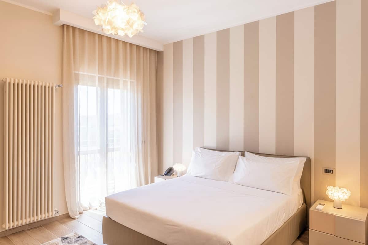 Quarto de hotel com uma cama grande, uma mesa de cabeceira com abajur, um radiador e uma janela com cortinas.