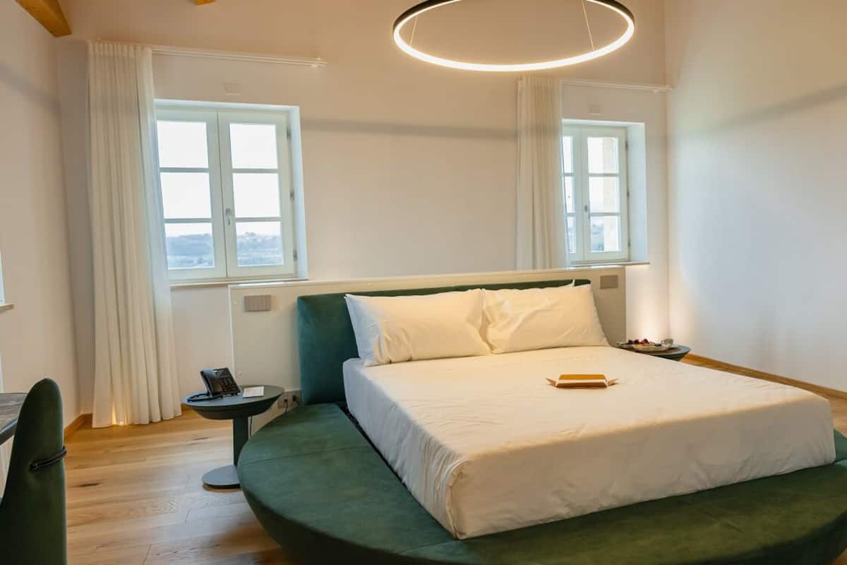 Quarto moderno com uma cama grande, mesa de cabeceira, cadeira e vista para o exterior pela janela.
