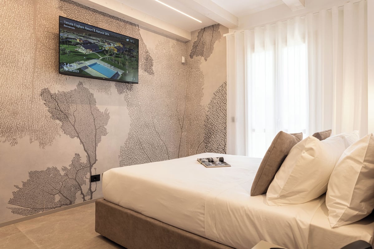 Quarto de hotel moderno com uma cama grande, uma TV de tela plana na parede e uma janela com cortinas leves.