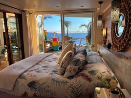 Quarto com uma cama grande, varanda com plantas em vasos e vista para o mar.