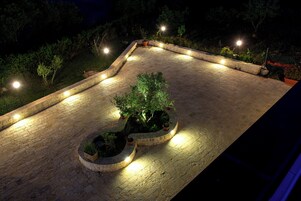 Uma área de estar ao ar livre com piso de pedra, plantas em vasos e várias luminárias.