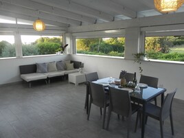 Um pátio amplo com mesa de jantar, cadeiras e um sofá. Janelas grandes oferecem vista para o verde.