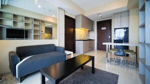 Uma sala de estar moderna com um sofá, uma mesa de centro e uma TV fixada na parede. Há uma área de cozinha com um refrigerador, uma pia e um balcão.