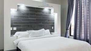 Quarto de hotel com design moderno, apresentando uma cama grande com roupa de cama branca, cabeceira em madeira escura e uma janela com cortinas.