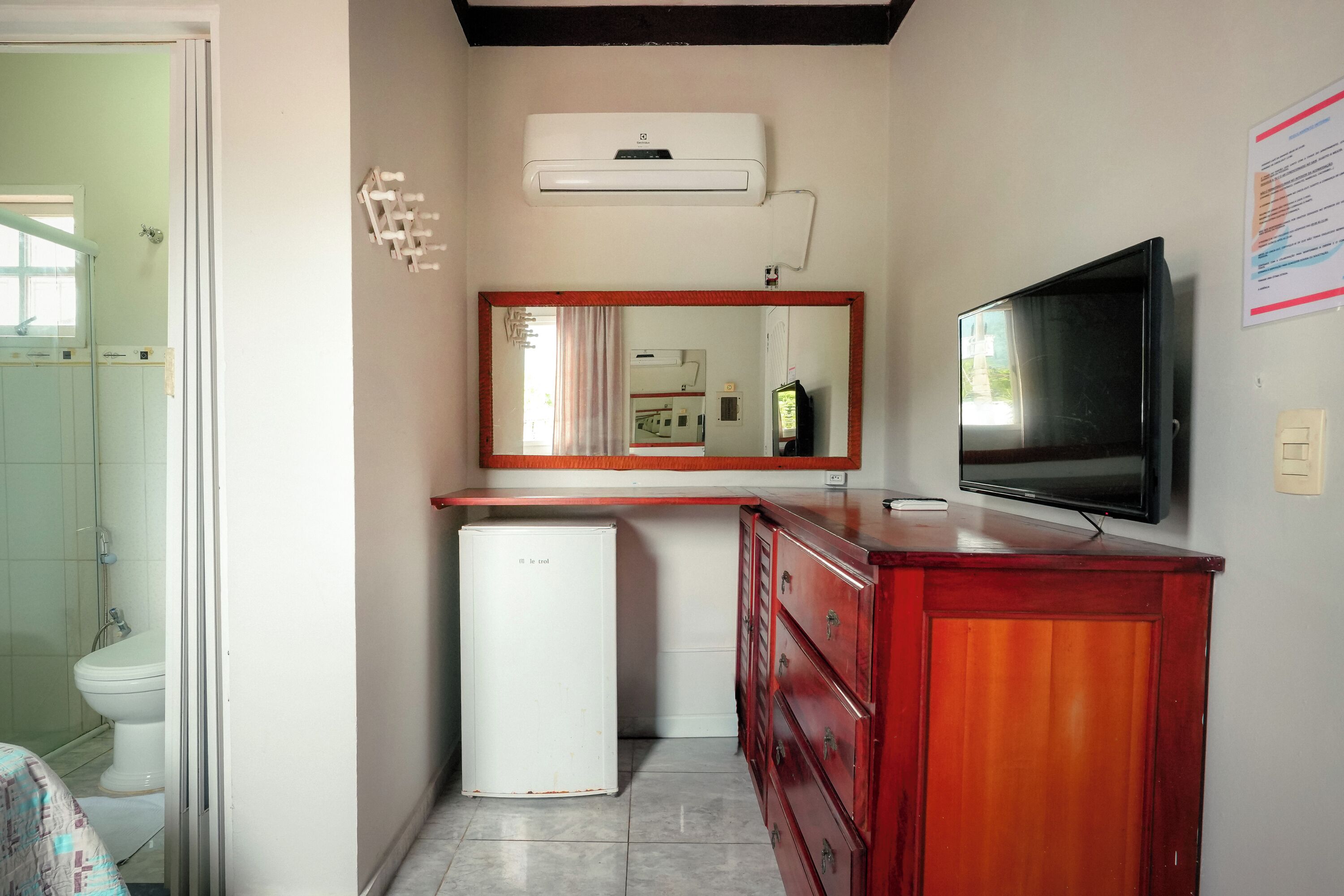 Uma casa de banho compacta com sanita branca, um frigorífico pequeno branco, toucador de madeira com espelho e uma televisão preta fixada na parede.