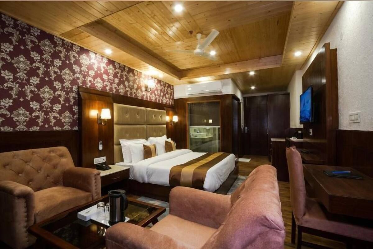 Quarto de hotel com teto de madeira, uma cama grande com cabeceira, duas poltronas, uma escrivaninha e uma televisão.