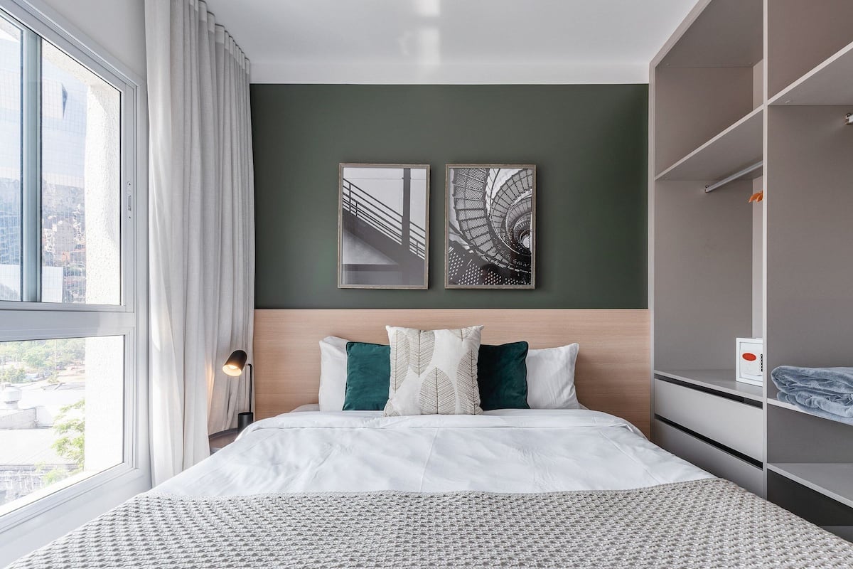 Quarto de hotel com cama, janela com cortinas, cabeceira e duas obras de arte penduradas na parede.