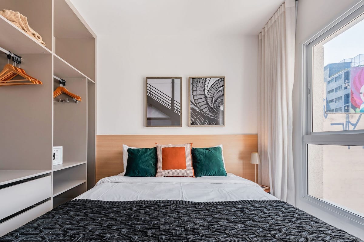 Quarto de hotel com uma cama, um guarda-roupa, duas imagens emolduradas e uma janela com vista.