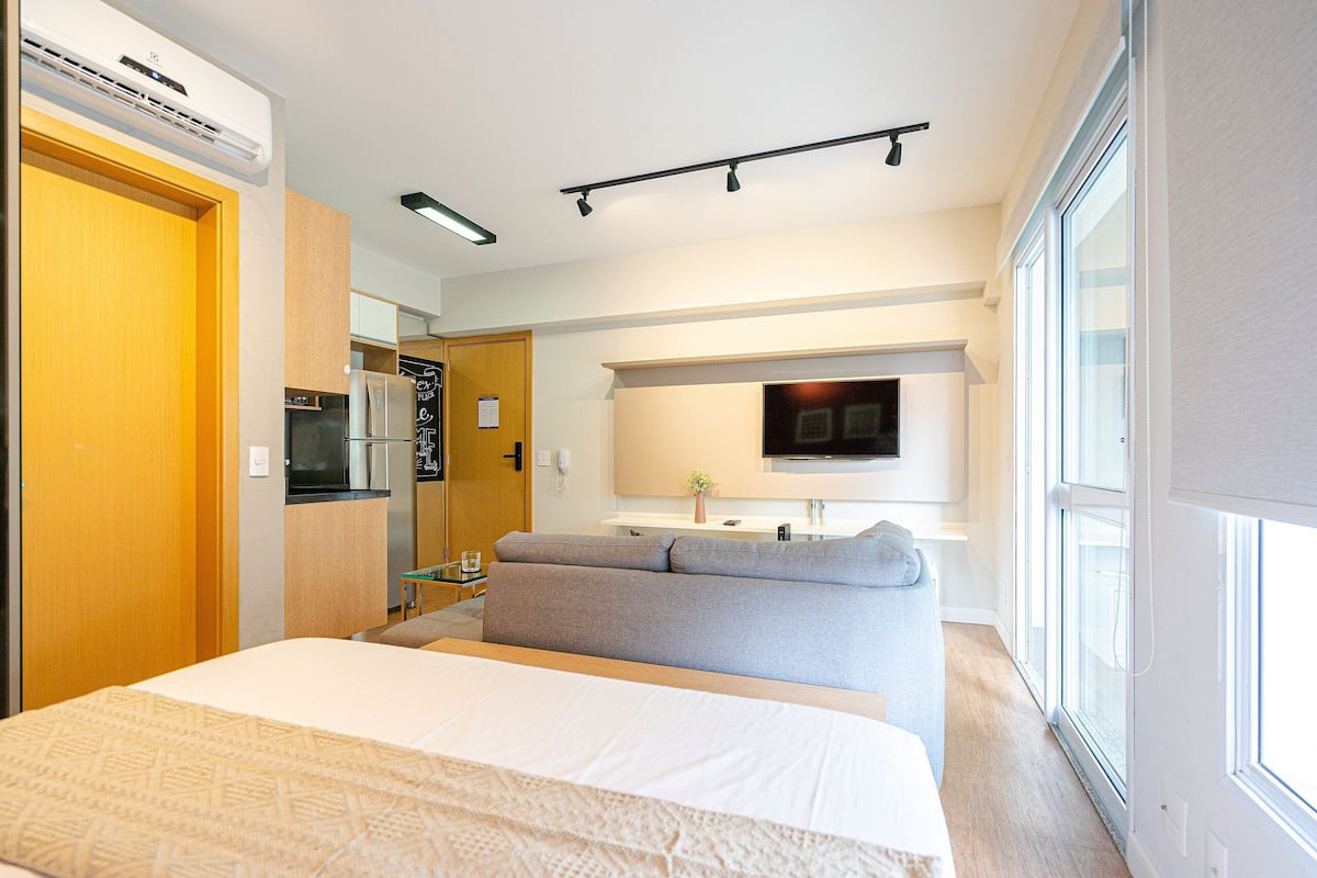 Quarto de hotel com cama, sofá, televisão, cozinha compacta e janela.