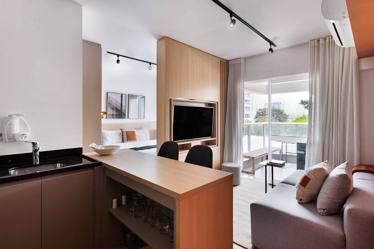 Quarto de hotel moderno com cozinha compacta, área de refeições, TV e varanda com vista.