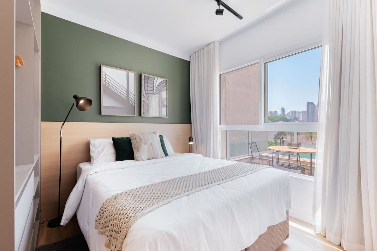 Quarto com uma cama grande, um abajur e vista para a cidade através de uma janela.