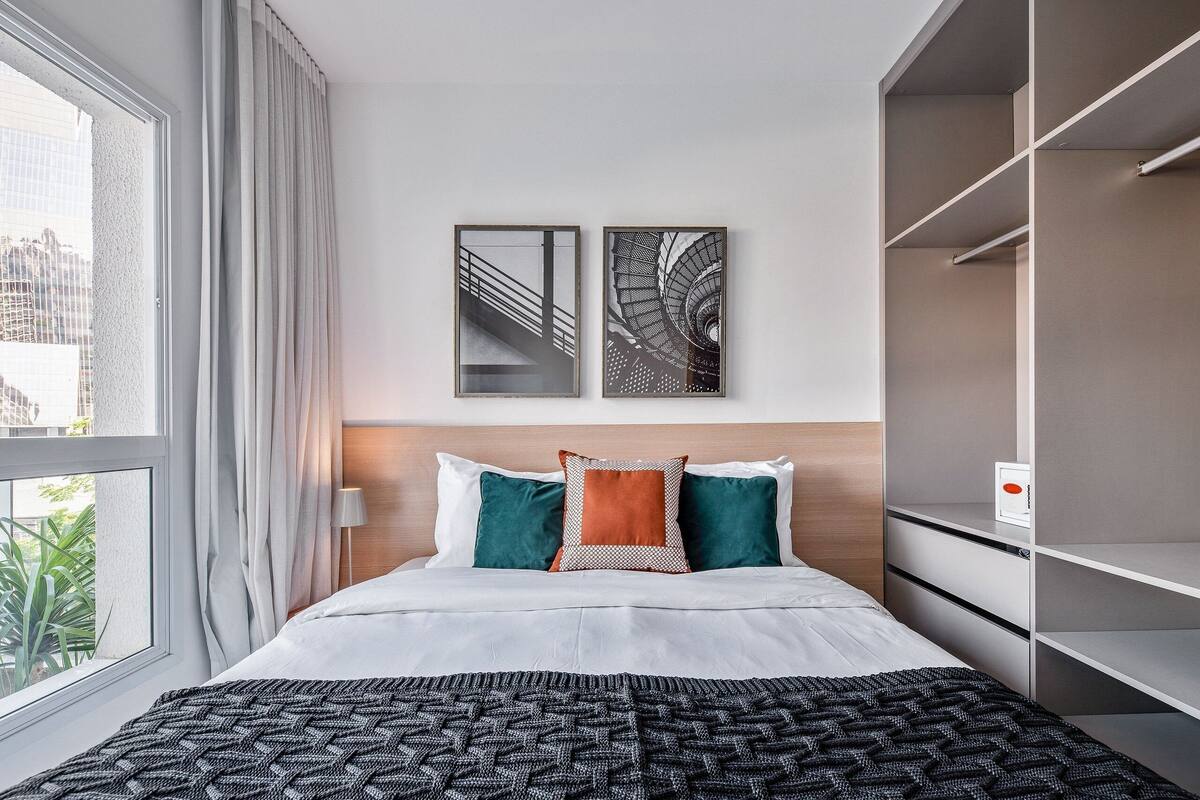 Quarto de hotel com cama, janela com cortinas, cabeceira e duas obras de arte fixadas na parede.