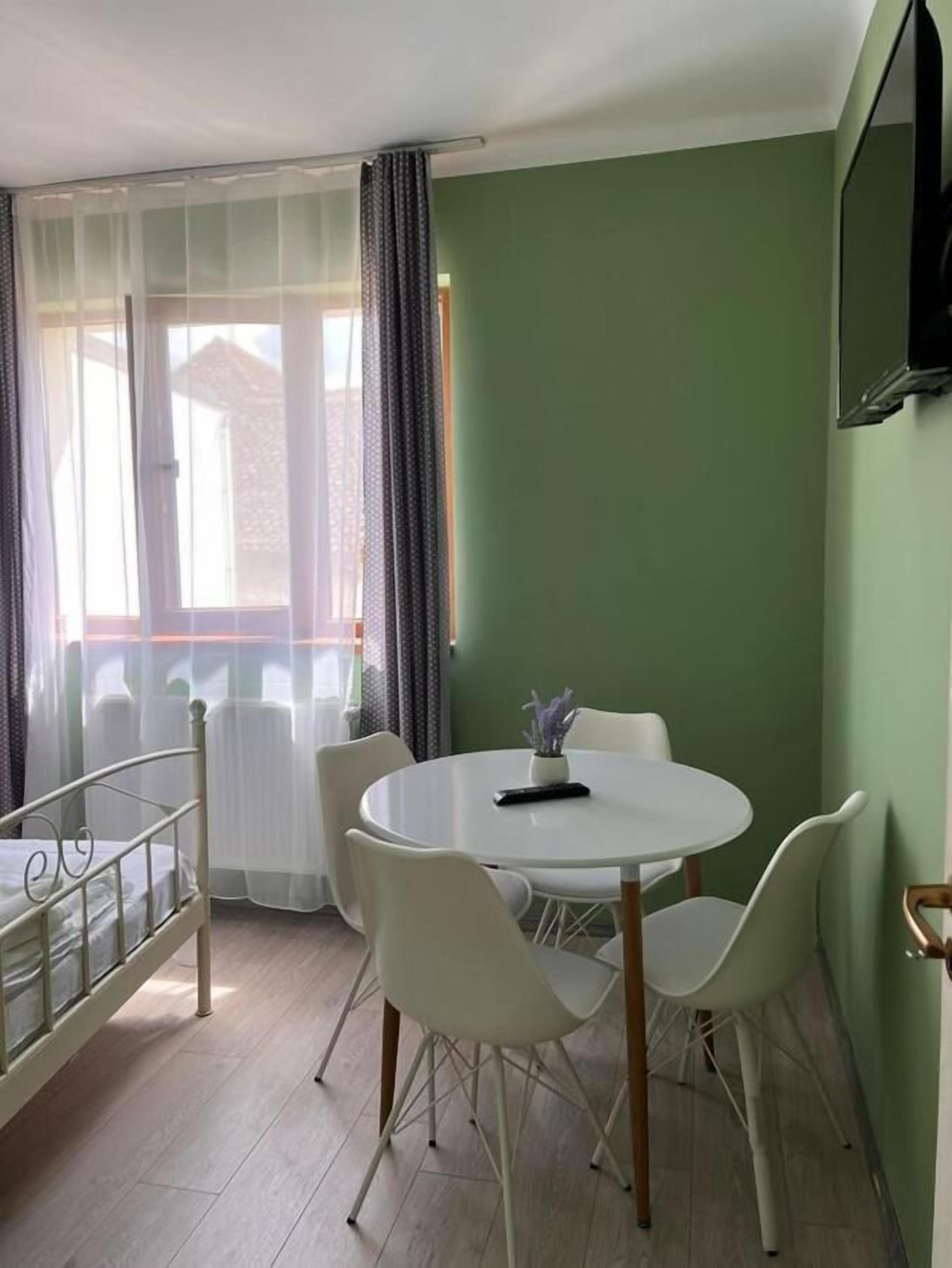 Quarto com uma mesa redonda, cadeiras brancas, uma cama com estrutura metálica e uma parede verde.