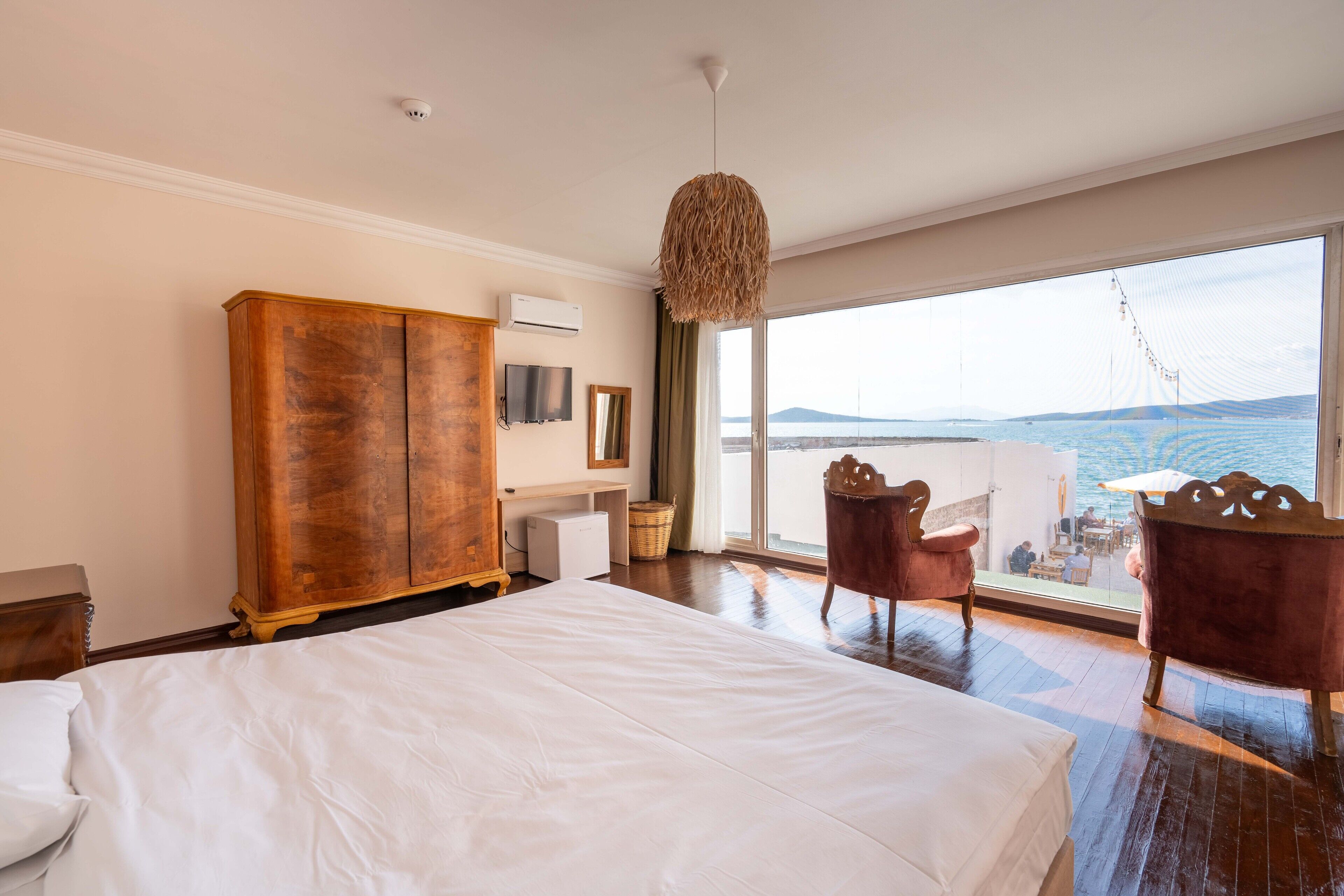 Quarto com cama grande, guarda-roupa de madeira e vista para o mar através de uma janela ampla.