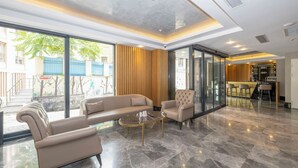 Um lobby moderno com sofá, poltrona e uma pequena mesa. Há um painel de vidro e uma área de recepção ao fundo.