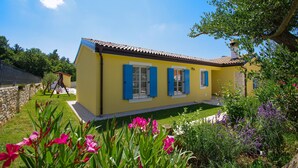 Uma casa amarela com persianas azuis, rodeada por um jardim bem cuidado com diversas plantas e flores.