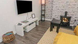 Uma sala de estar moderna com uma TV de tela plana, um móvel branco para equipamentos eletrônicos, uma estante, uma cesta de fibra natural, um cobertor amarelo, um tapete estampado e uma lareira a lenha.