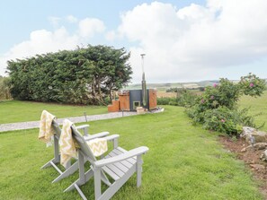 Uma área externa gramada com duas cadeiras Adirondack, um deck de madeira e um pequeno edifício com chaminé.