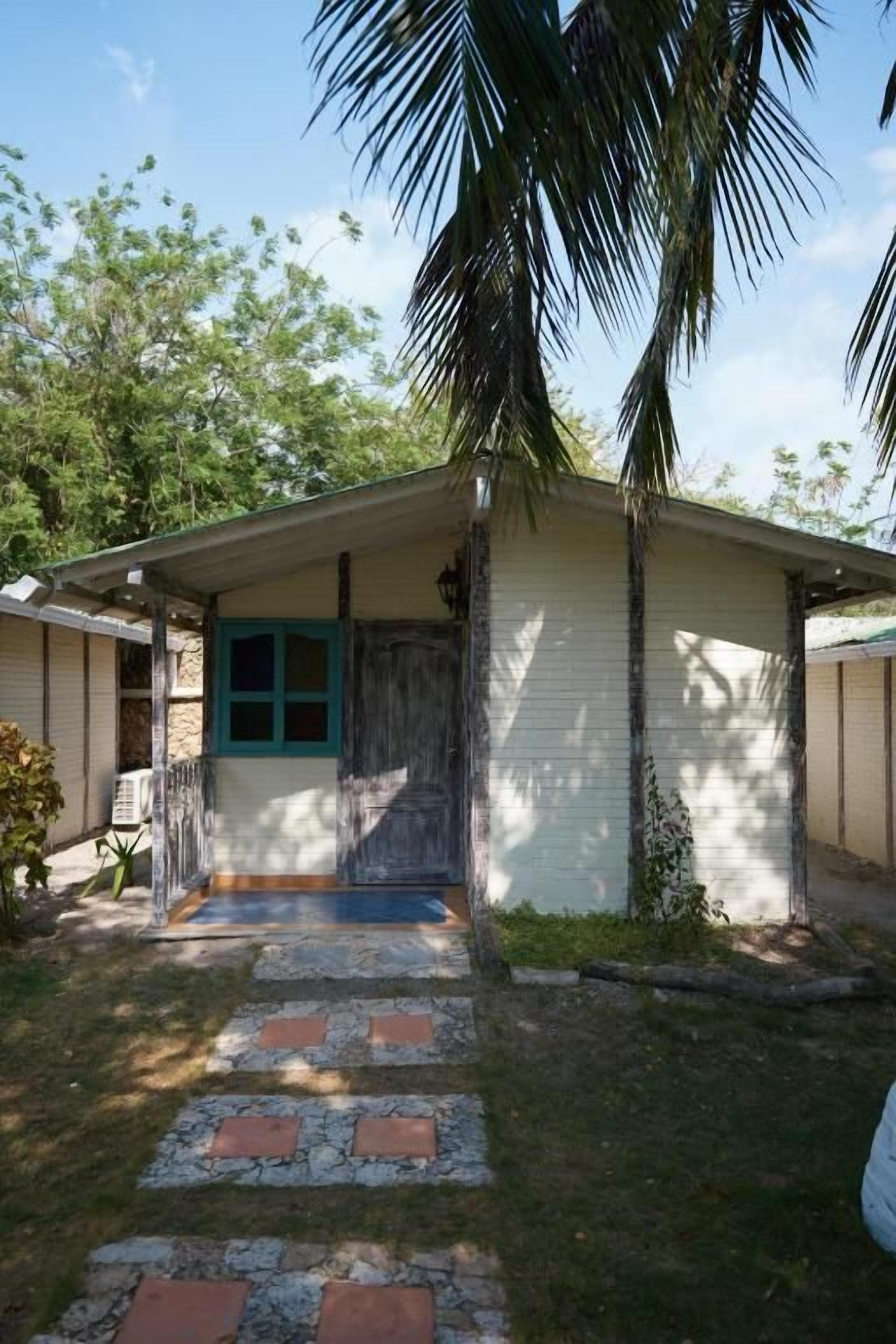 Uma casa térrea com varanda frontal, porta com janela e pequeno jardim da frente com uma palmeira.