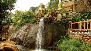 Uma cascata deságua em um lago, rodeado por vegetação exuberante e um muro de pedra com corrimão.