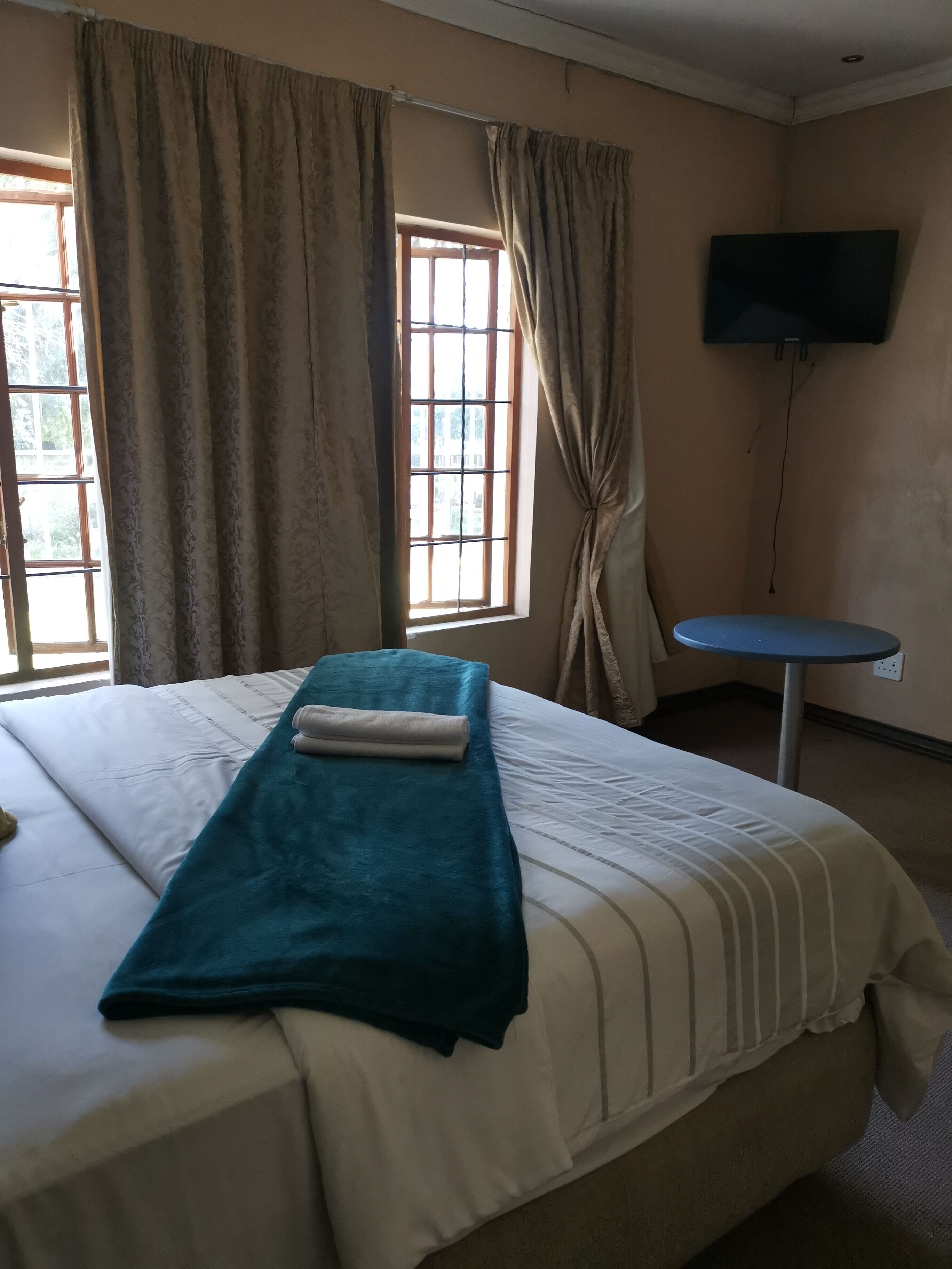 Quarto de hotel com uma cama, uma mesinha redonda, uma TV fixada na parede e uma janela com cortinas.