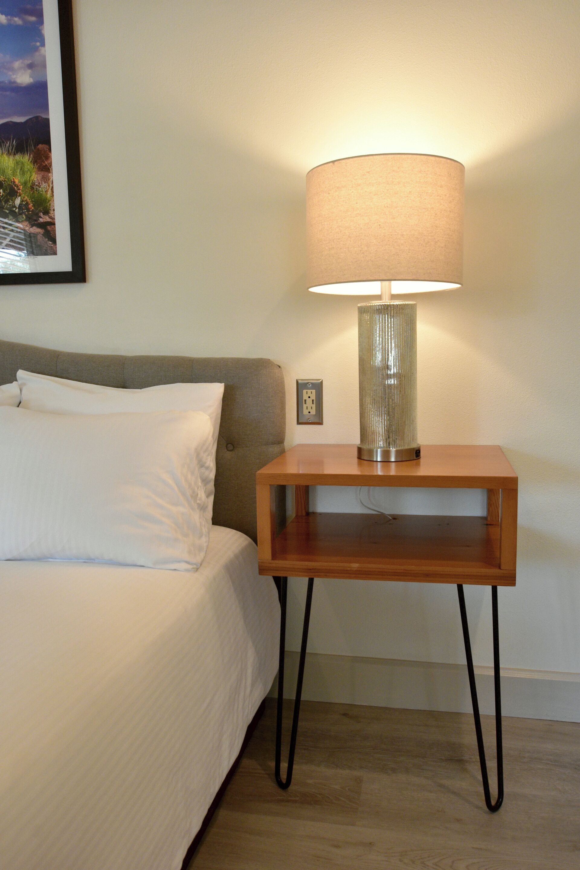 Uma cama bem arrumada com roupa de cama branca, uma mesa de cabeceira de madeira com abajur e um quadro na parede.