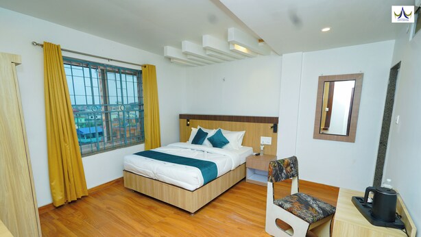 Quarto de hotel com piso de madeira, uma cama com cabeceira de madeira, uma cadeira, mesa de cabeceira com chaleira e uma janela com cortinas.