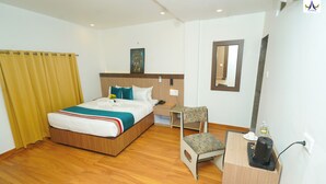 Quarto de hotel com piso de madeira, uma cama com colcha branca e azul-turquesa, uma pequena escrivaninha com cadeira e um espelho na parede.