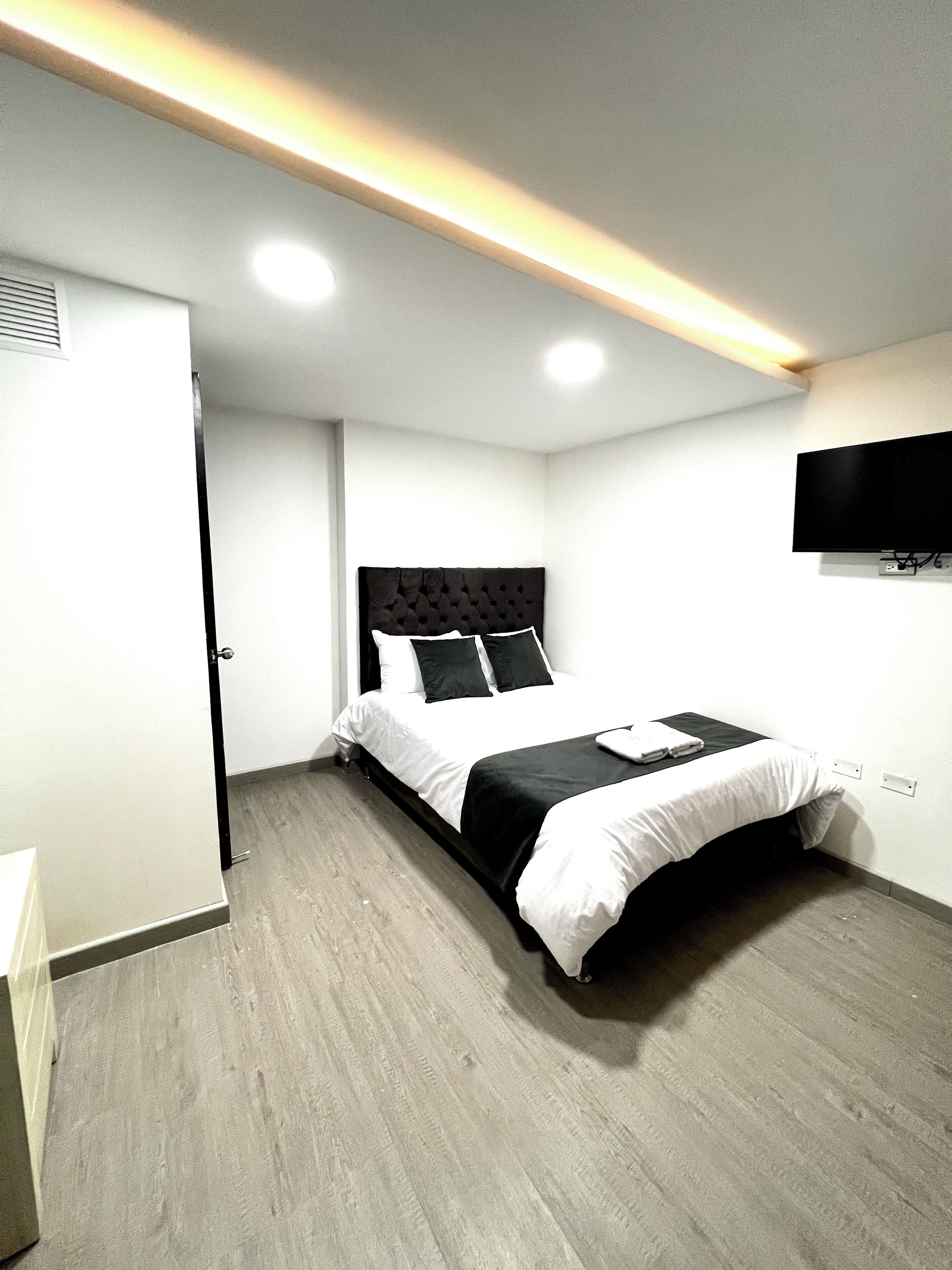 Uma cama de solteiro com cabeceira preta e roupa de cama branca em um quarto com piso de madeira e paredes brancas.