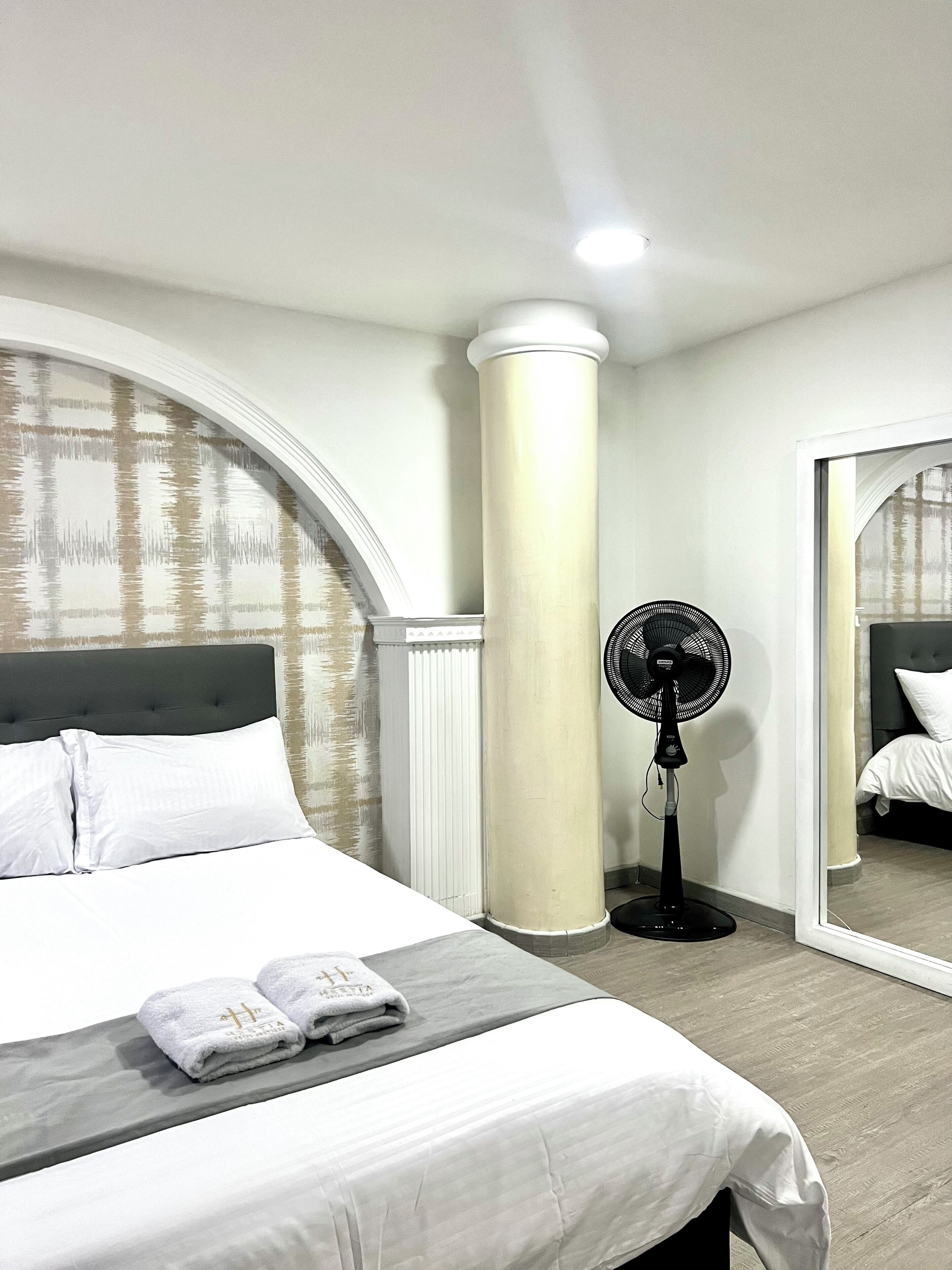 Quarto de hotel com uma cama, um ventilador, um espelho grande e um pilar branco.