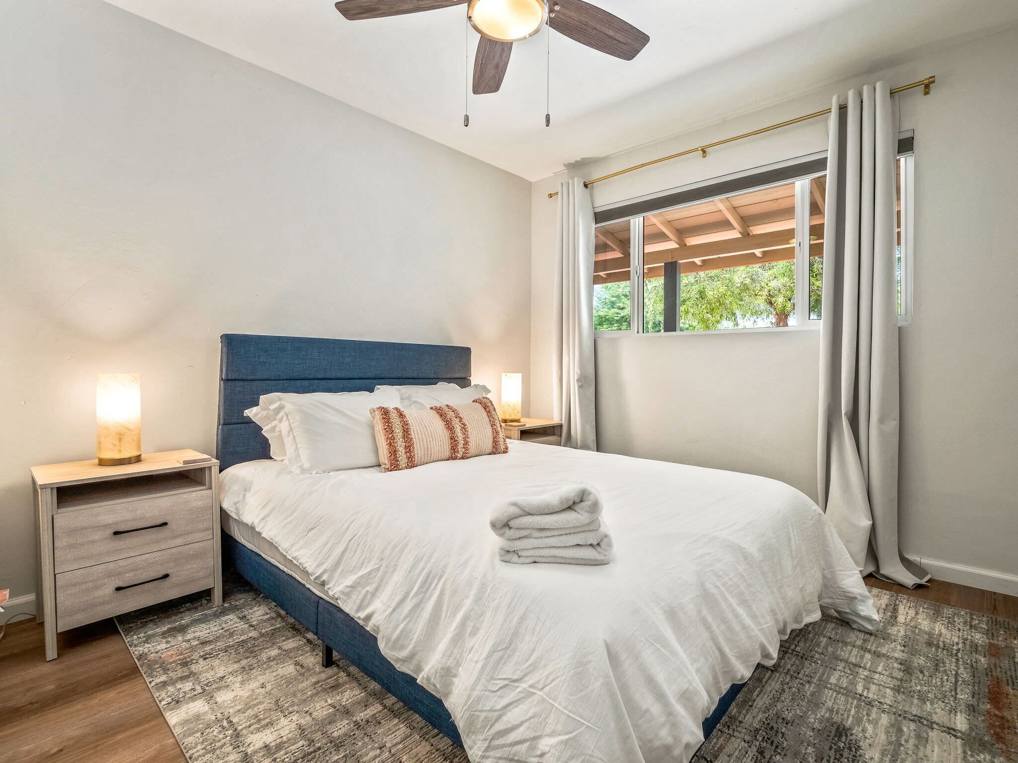 Quarto com cama, mesas de cabeceira, ventilador de teto e janela com cortinas.