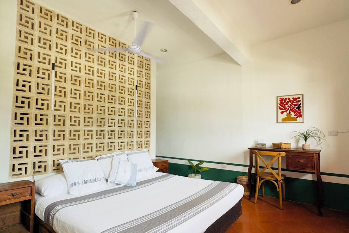 Um quarto com uma cama, uma escrivaninha, uma cadeira, um painel decorativo na parede e um ventilador de teto.