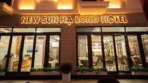 Exterior do Hotel New Sun Ha Long, com letreiros iluminados, grandes portas de vidro e uma área de recepção visível no interior.
