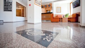 Recepção de hotel com piso espelhado, balcão de atendimento, área de estar e televisão fixada na parede.