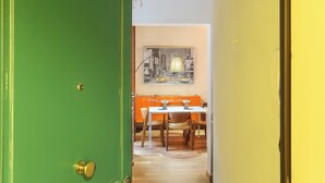 Uma porta verde dá acesso a uma sala de jantar com mesa e cadeiras, um quadro com vista urbana enquadrada na parede e um lustre.