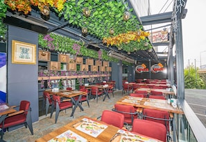 Restaurante interno com cadeiras vermelhas, mesas de madeira e um teto decorado com plantas e luminárias pendentes.