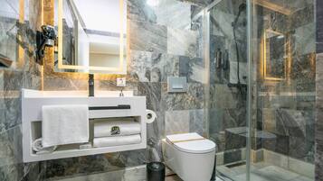 Uma casa de banho moderna com chuveiro em cabine de vidro, paredes de mármore, toucador branco com barra de toalhas e sanita.