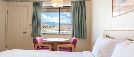 Quarto de hotel com cama, mesa com cadeiras, janela com cortinas e vista para uma paisagem.