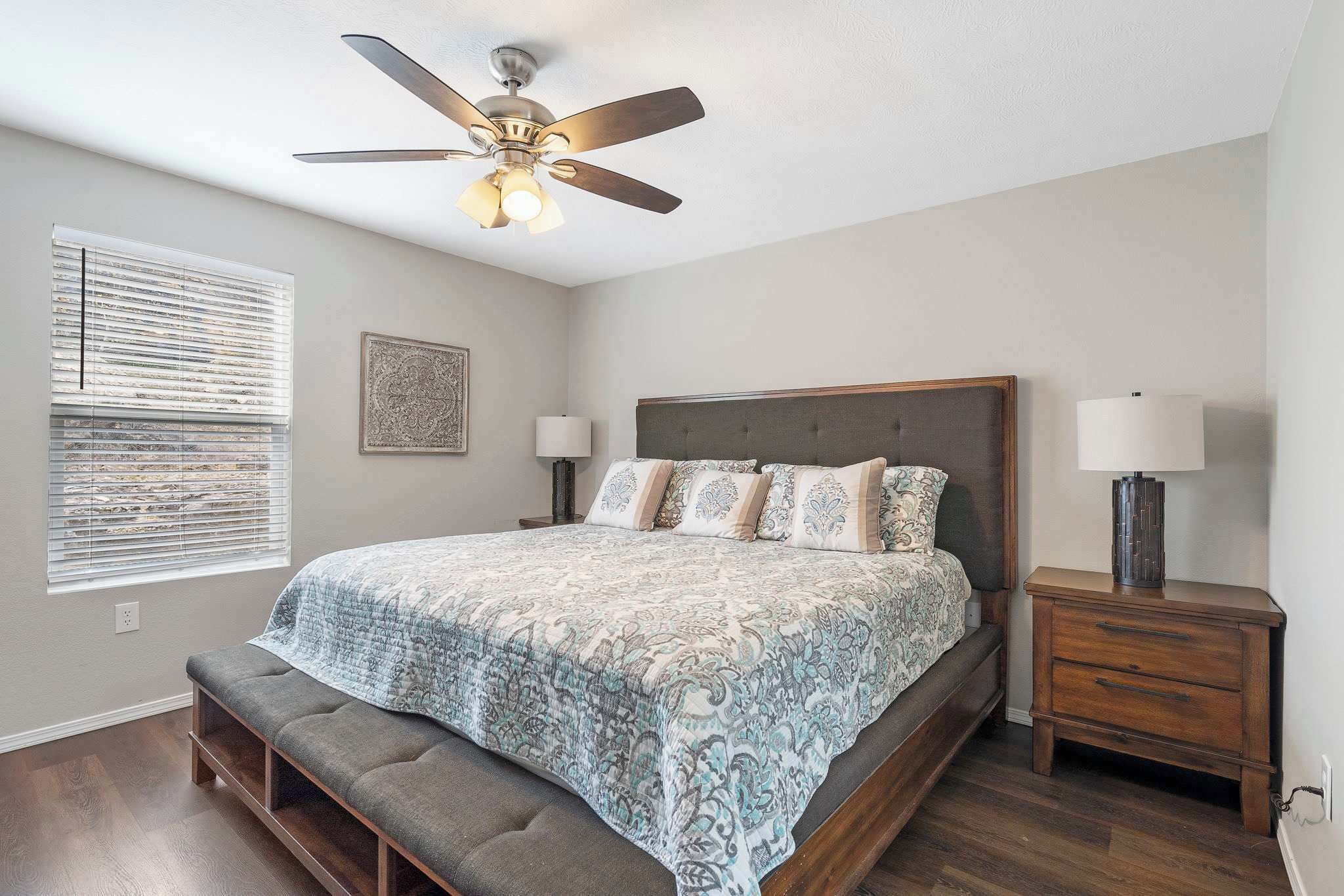 Quarto com cama, um banco, um ventilador de teto, duas mesas de cabeceira e uma janela com persianas.