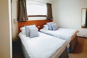 Quarto de hotel com duas camas de solteiro, cabeceira de madeira, janela com cortinas e um espelho na parede, além de piso coberto por carpete.