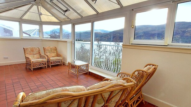 Uma sala de sol com móveis de vime, janelas amplas e vista para montanhas.