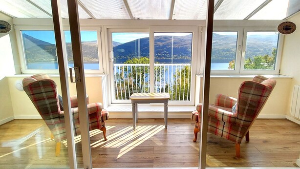 Quarto com vista para um lago e montanhas, cadeiras xadrez, uma mesa e um painel de vidro.