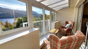Varanda com vista para um lago e montanhas, mobiliada com cadeiras xadrez e uma mesa pequena.