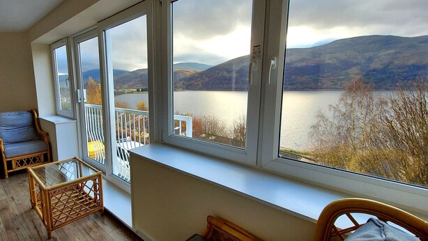 Uma varanda com móveis de vime, uma mesa de vidro e vista para um lago e montanhas.