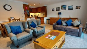 Sala de estar com dois poltronas, um sofá, uma mesa de centro e um espaço de jantar com mesa e cadeiras. Cozinha ao fundo, com armários de madeira e um relógio na parede.